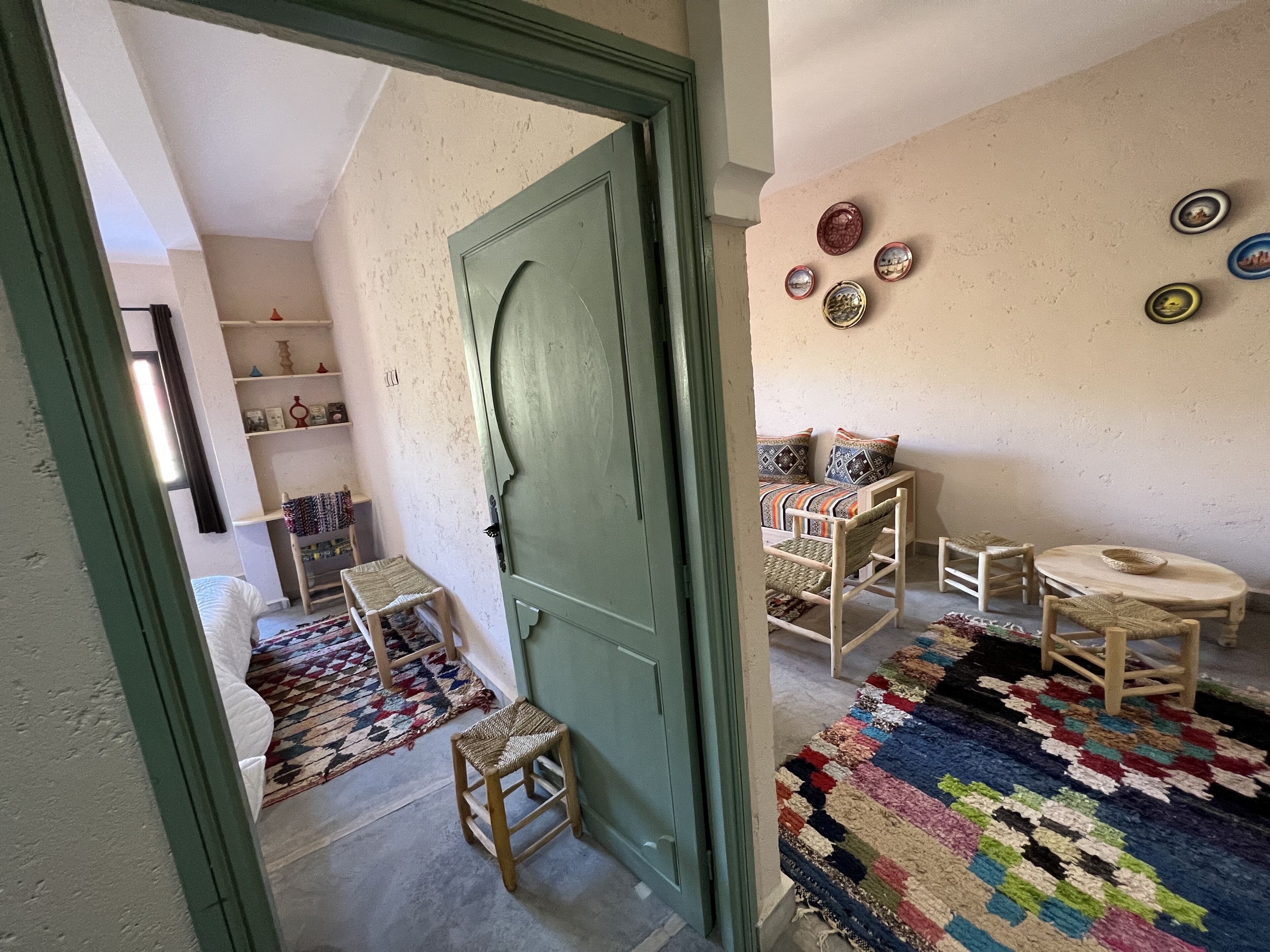 Um quarto aconchegante com uma porta verde, um banquinho de madeira, uma cadeira de fibra natural, um tapete estampado e uma parede decorada com placas de metal.