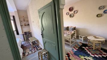 Um quarto aconchegante com uma porta verde, um banquinho de madeira, uma cadeira de fibra natural, um tapete estampado e uma parede decorada com placas de metal.