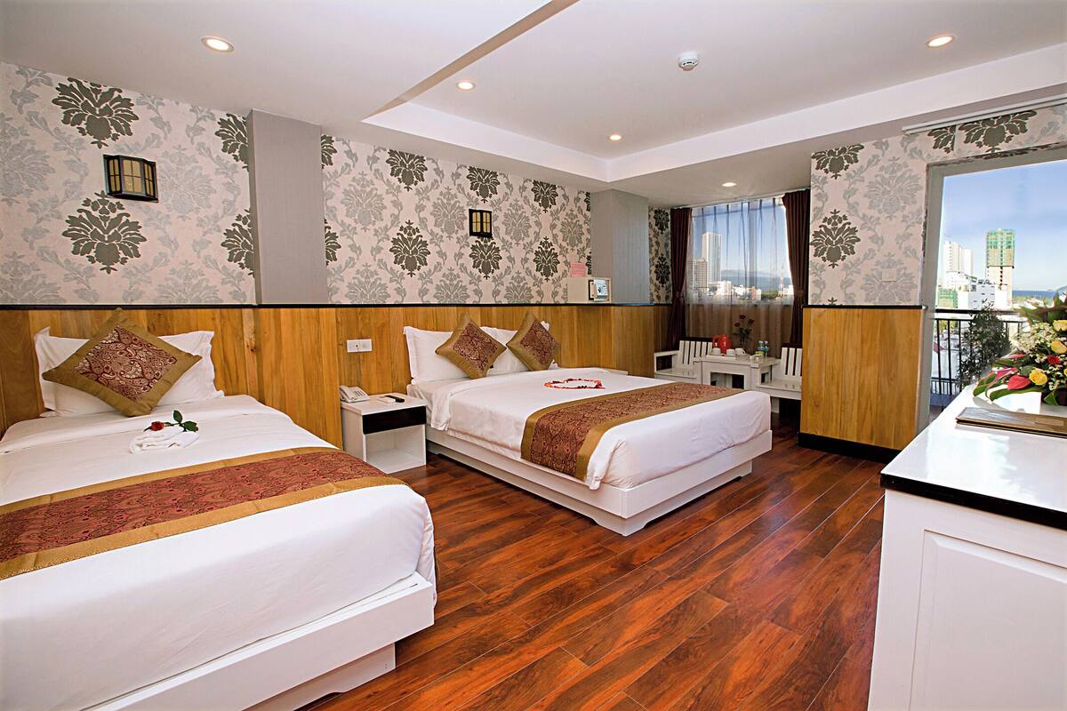 Quarto de hotel com duas camas, painéis de madeira, papel de parede floral, uma escrivaninha com um vaso de flores e vista para a cidade através de grandes janelas.