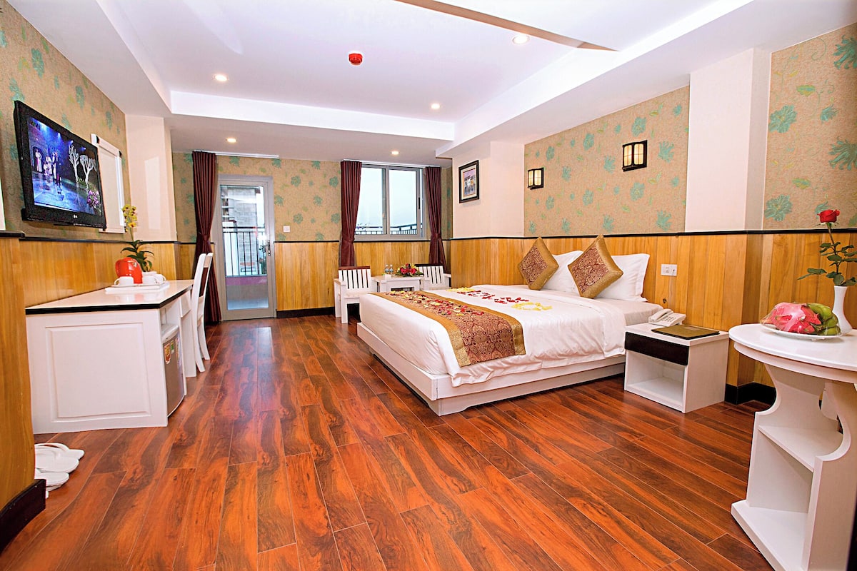 Quarto de hotel com cama, mesa de cabeceira, escrivaninha, cadeira e televisão na parede.