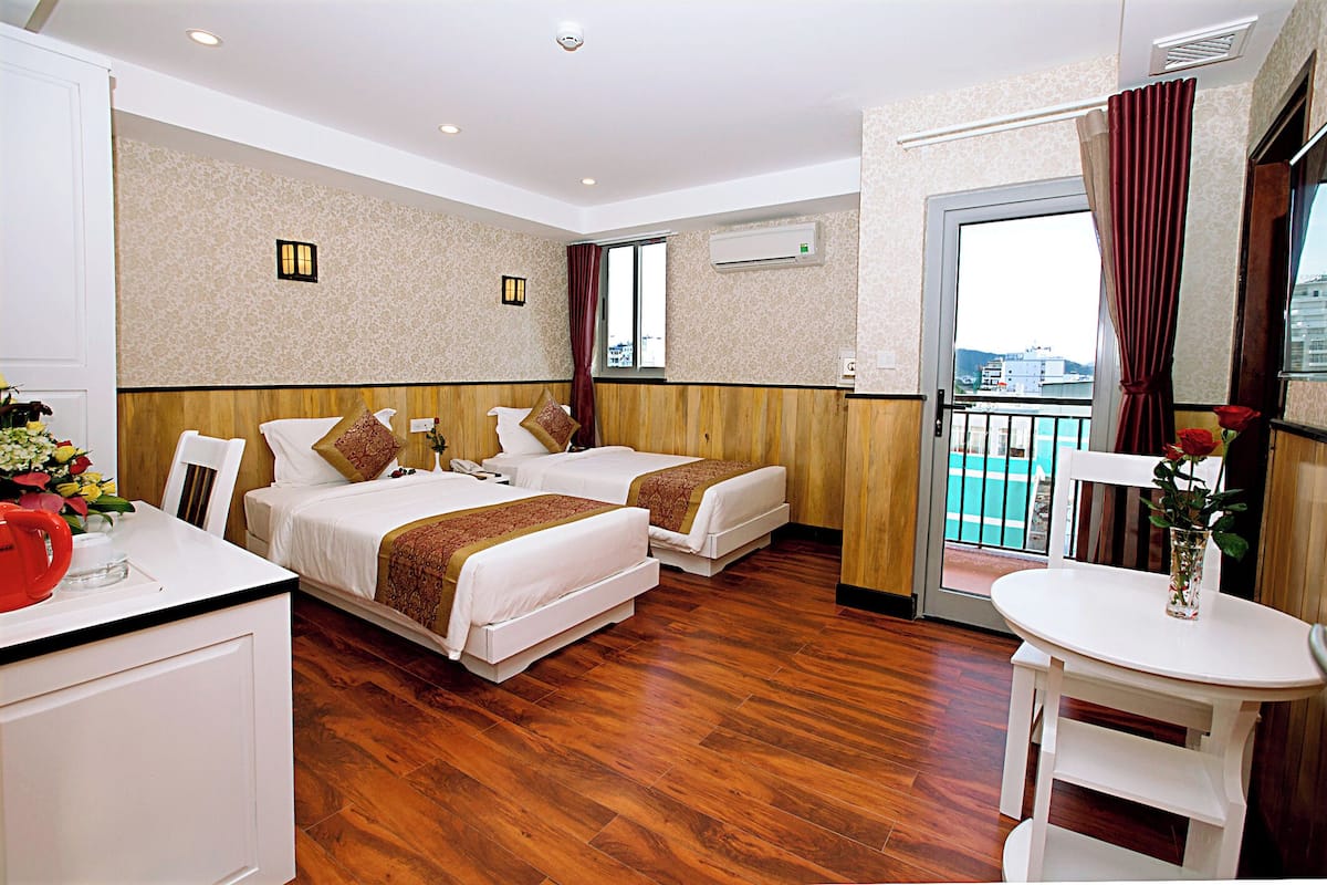 Quarto de hotel com duas camas, uma mesinha, uma cadeira, uma escrivaninha e vista para o exterior.