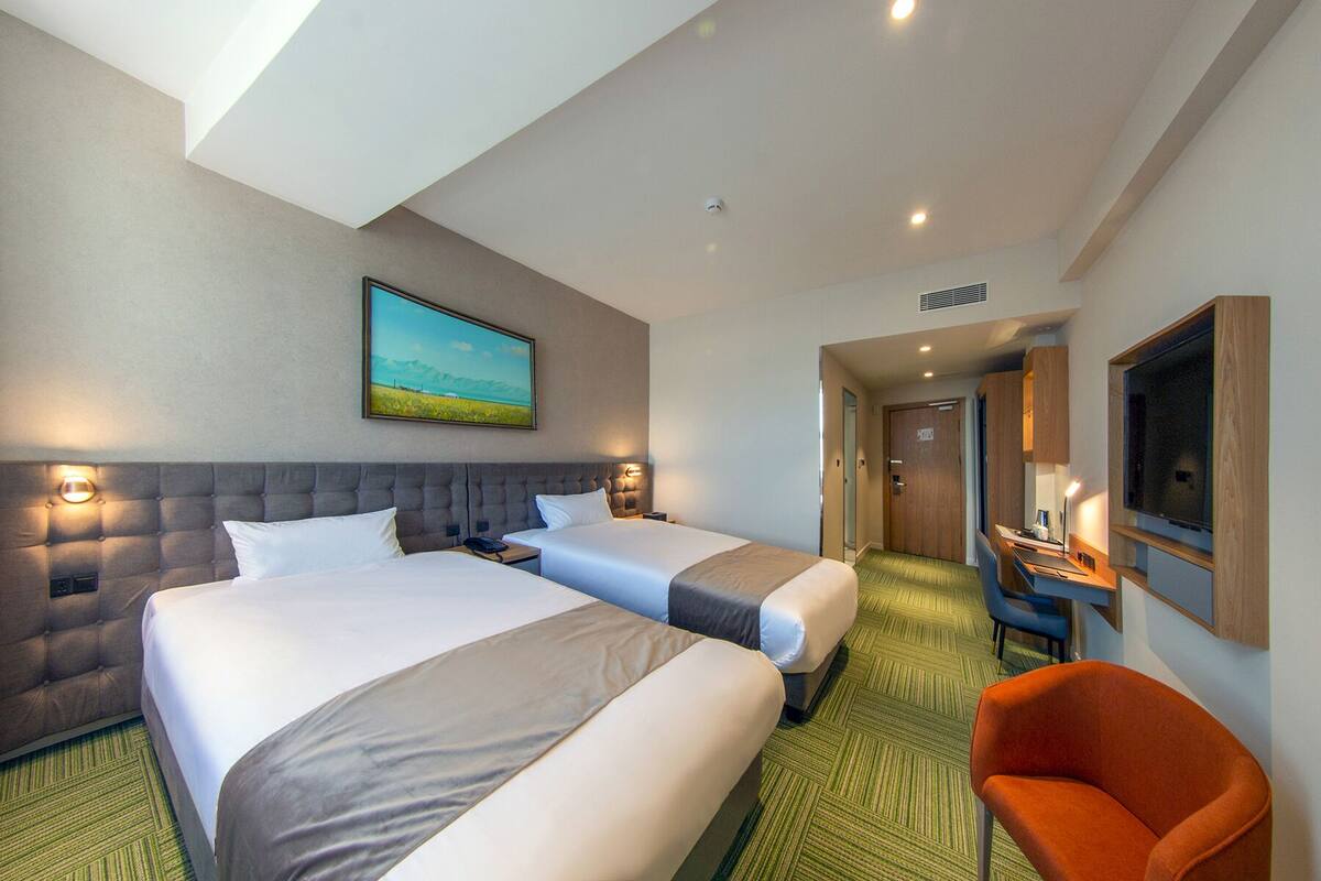 Quarto de hotel com duas camas, uma janela grande, um quadro na parede e uma mesa com uma cadeira.