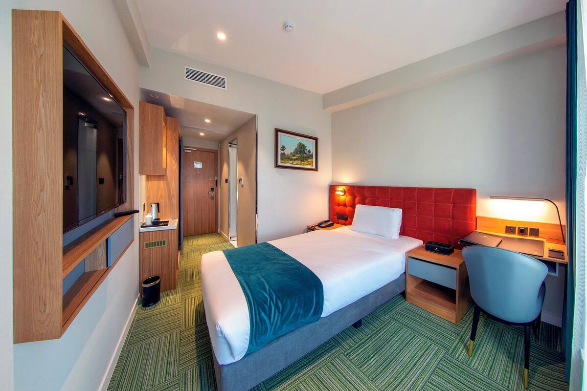 Quarto de hotel com cama, escrivaninha, cadeira e televisão.