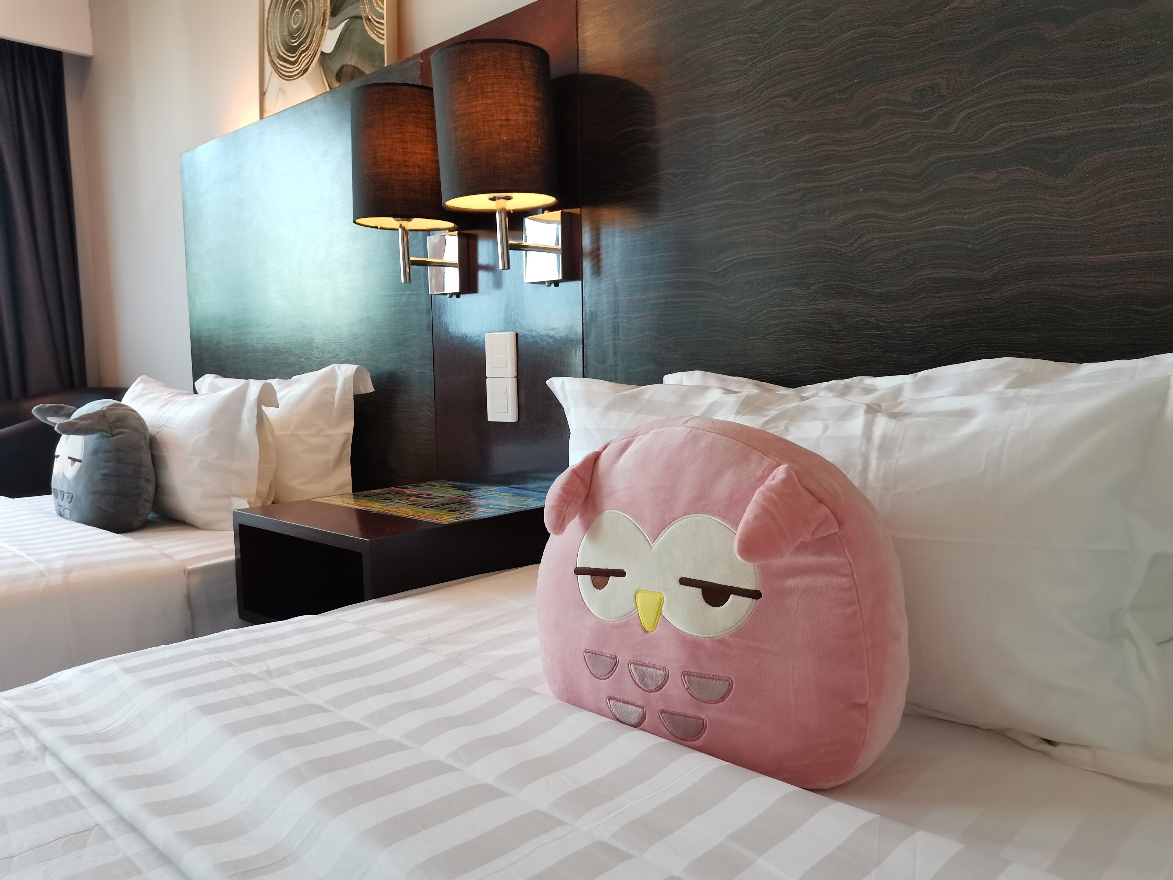Quarto de hotel com cama, almofadas, mesa de cabeceira e luminária fixada na parede.