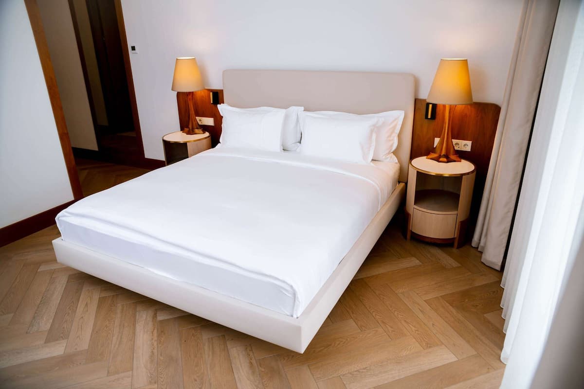 Cama bem arrumada com lençóis brancos, duas mesas de cabeceira com luminárias e piso de madeira em padrão herringbone.