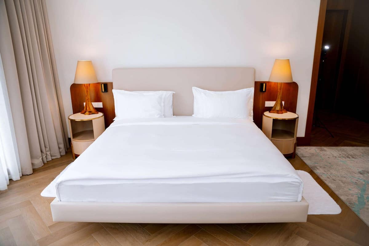 Uma cama grande com roupa de cama branca, duas mesas de cabeceira com luminárias e um piso de madeira.