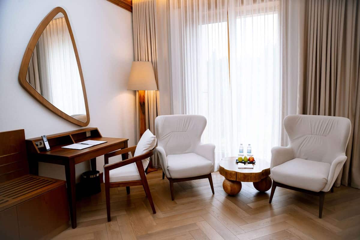 Quarto de hotel com escrivaninha, duas cadeiras, mesa de madeira redonda com frutas, luminária e um espelho grande.