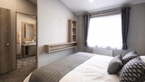 Quarto com cama, guarda-roupa, prateleira embutida, janela com cortinas e banheiro visível por uma porta aberta.