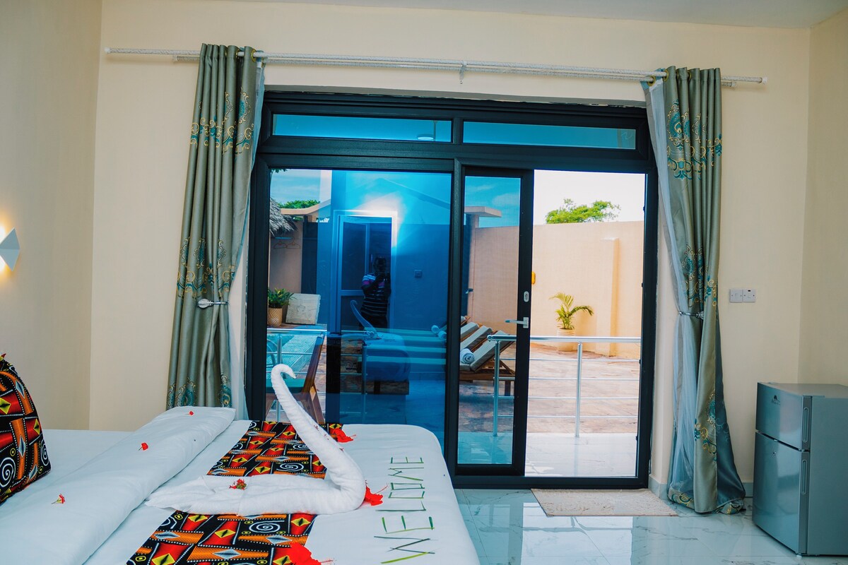 Quarto de hotel com cama, tapete de boas-vindas, geladeira e uma porta de vidro deslizante para uma área externa com mesa e cadeiras.