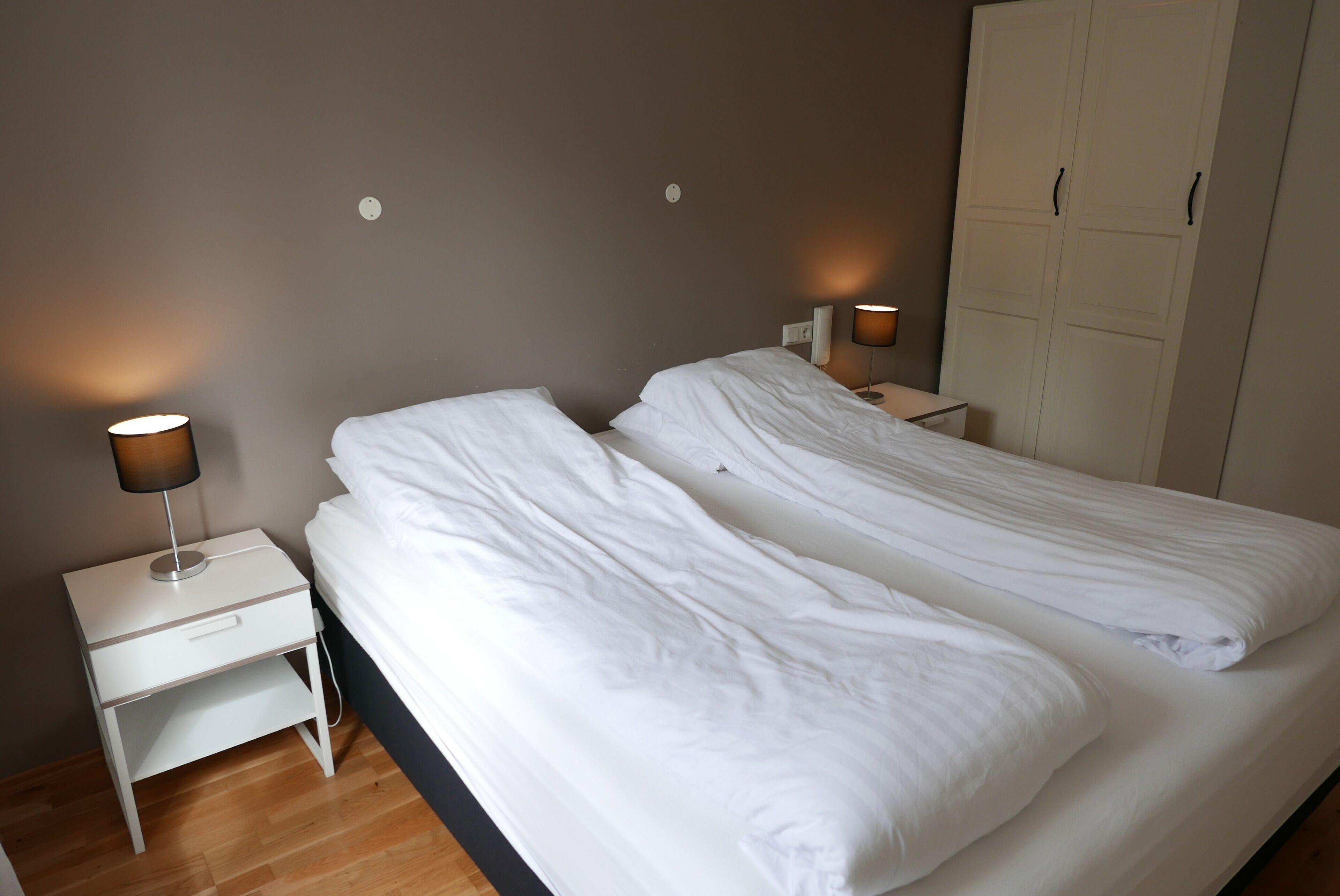 Uma cama de casal com roupa de cama branca, uma mesa de cabeceira com abajur e um guarda-roupa em um quarto de hotel.
