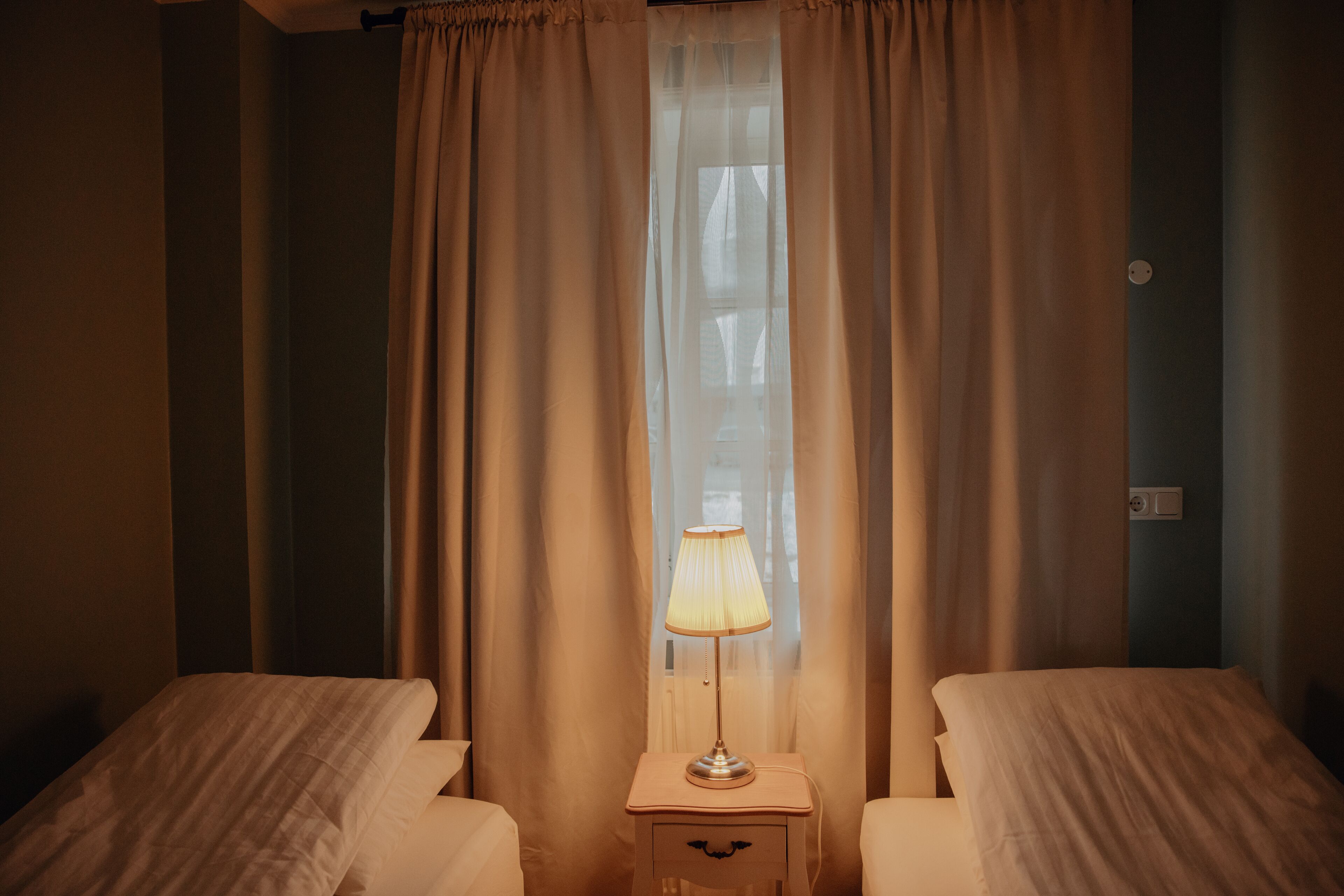 Quarto de hotel com duas camas, uma mesa de cabeceira com abajur e uma janela com cortinas.