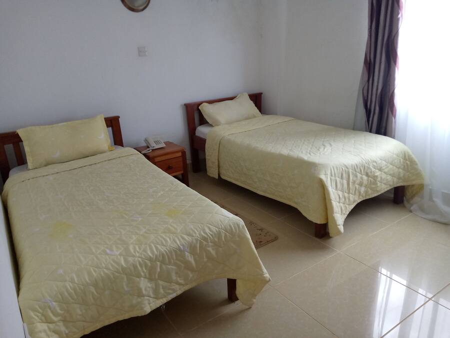 Duas camas de solteiro com roupa de cama amarela combinando, uma mesa de cabeceira de madeira com telefone e piso de azulejos.
