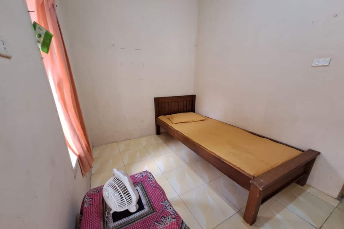 Quarto pequeno e simples com uma cama de solteiro, um ventilador e um tapete estampado.