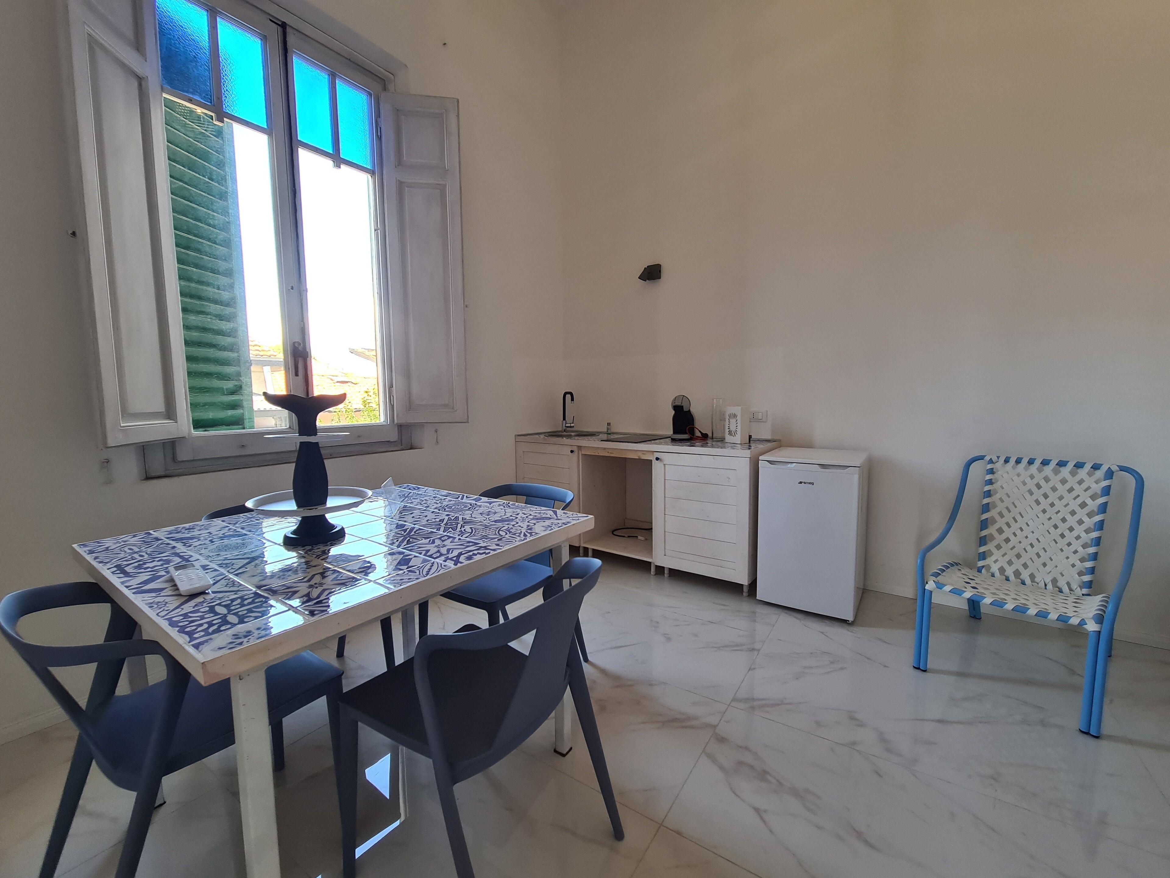 Quarto pequeno e limpo com soalho em mosaico, uma mesa com cadeiras, uma kitchenette compacta e uma cadeira azul.