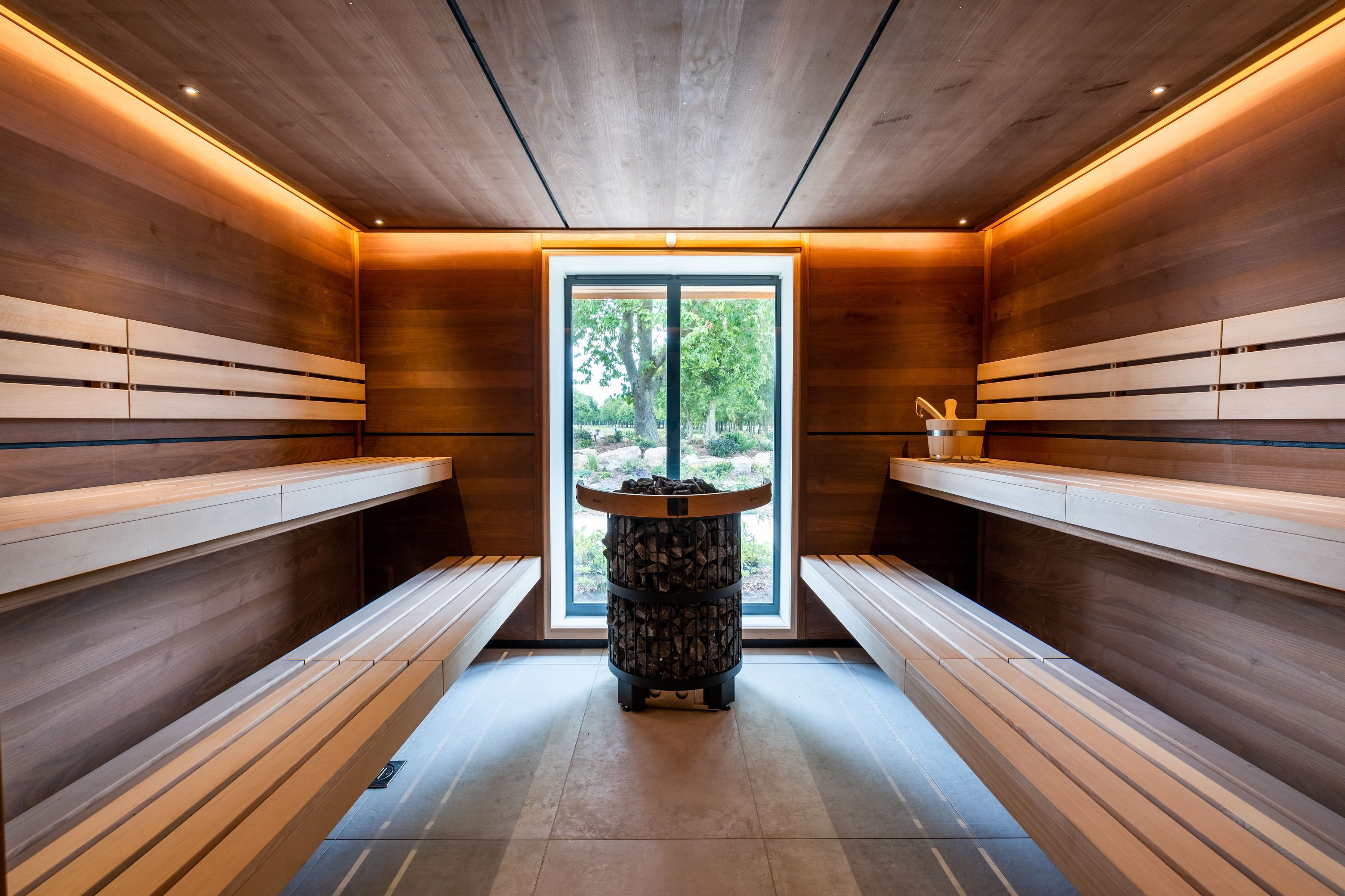 Uma sala de sauna com bancos de madeira, uma fogueira central e uma grande janela com vista para o exterior.