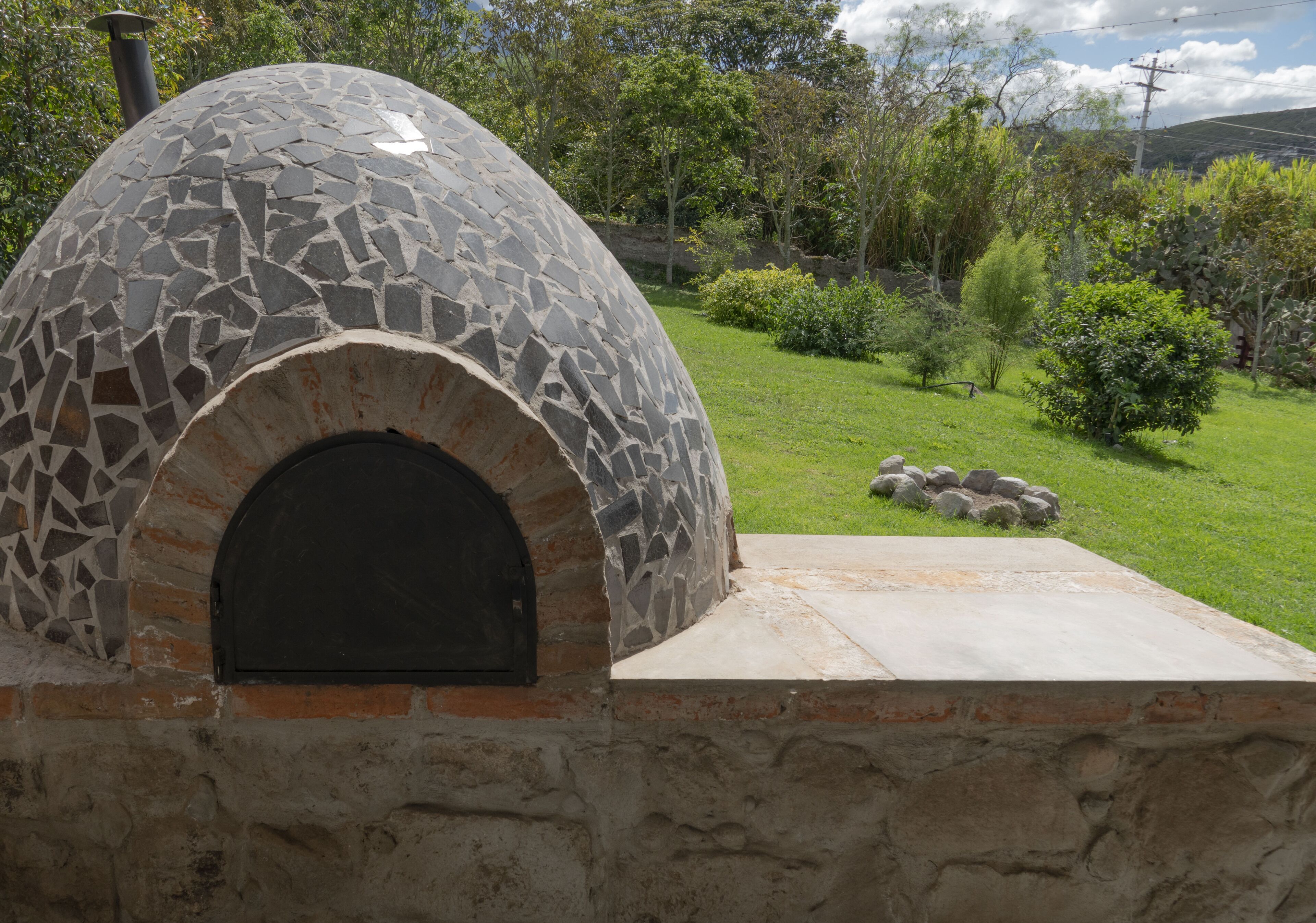 Forno de pedra com cobertura em cúpula de mosaico, situado numa área gramada com árvores e rochas ao fundo.