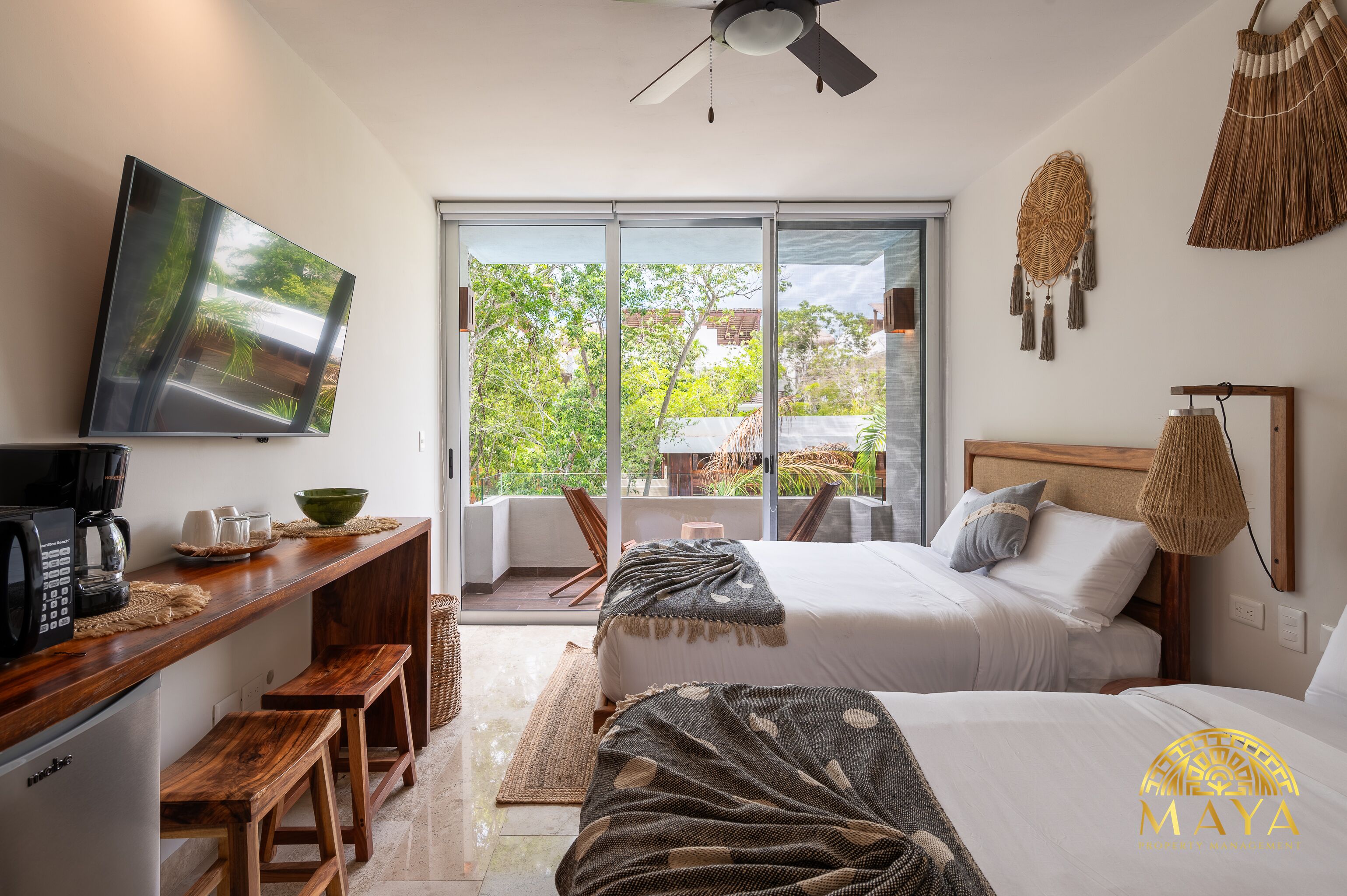 Quarto de hotel moderno com uma televisão grande, uma mesa de madeira com máquina de café, duas camas e uma varanda com vista para áreas verdes.