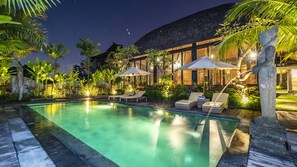 Uma área de piscina à noite, com espreguiçadeiras e guarda-sóis, rodeada por plantas tropicais e um edifício com arquitetura de madeira.