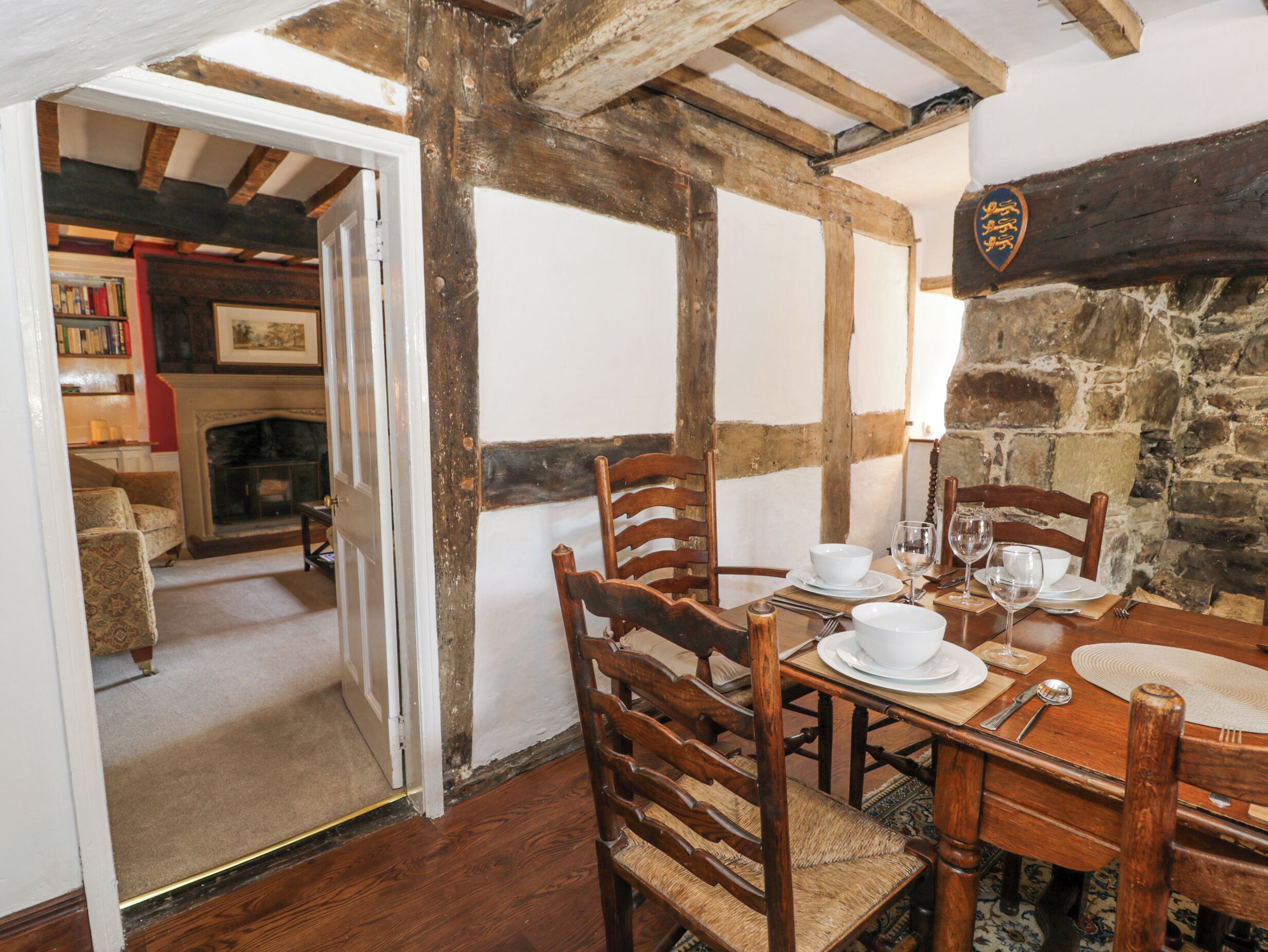 Uma sala de jantar tradicional com uma mesa de madeira posta para quatro pessoas, rodeada por cadeiras de madeira. Uma parede de pedra e uma lareira são visíveis ao fundo.