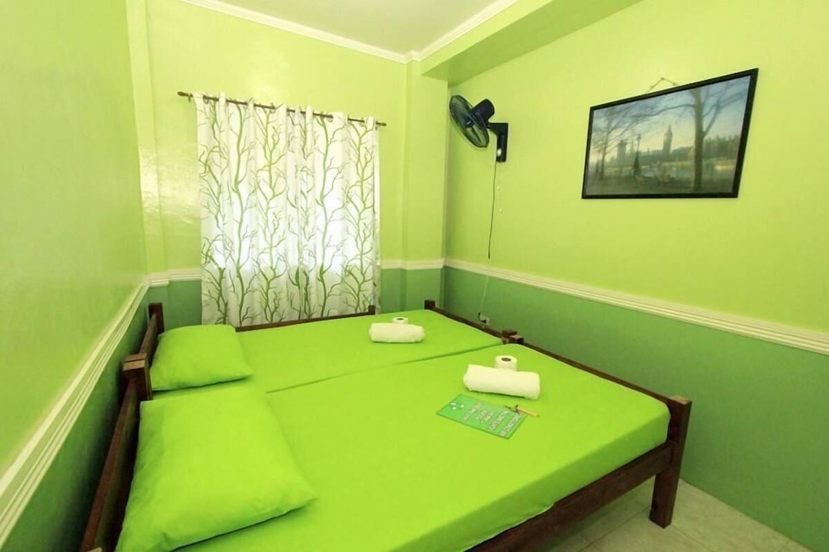 Quarto com paredes verdes, cama com roupa de cama verde, janela com cortina estampada com padrão de árvore, ventilador fixado na parede e um quadro emoldurado.