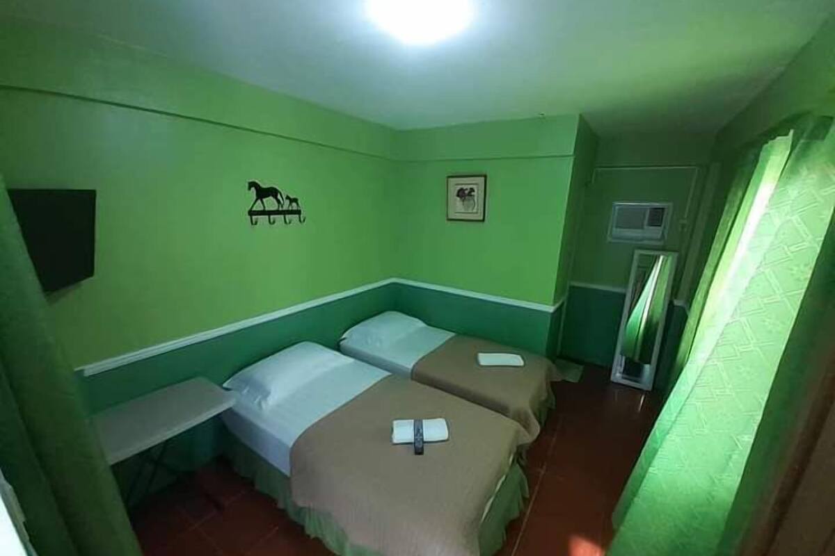 Quarto pequeno com decoração em tons de verde, duas camas de solteiro, uma mesinha e ar condicionado na parede.