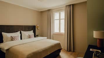 Quarto de hotel com uma cama grande, uma escrivaninha com candeeiro, janela com cortinas e um relógio fixado na parede.