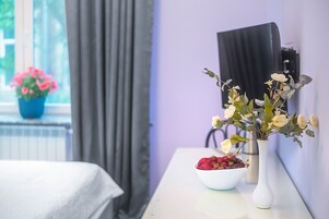 Quarto com uma mesa branca, um vaso branco com flores, uma tigela de frutas e uma janela com cortinas.
