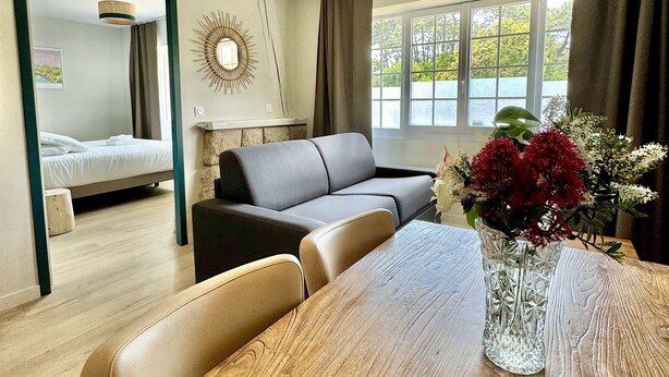 Uma sala de estar moderna com uma mesa de jantar de madeira, um sofá cinza e uma janela grande com cortinas.