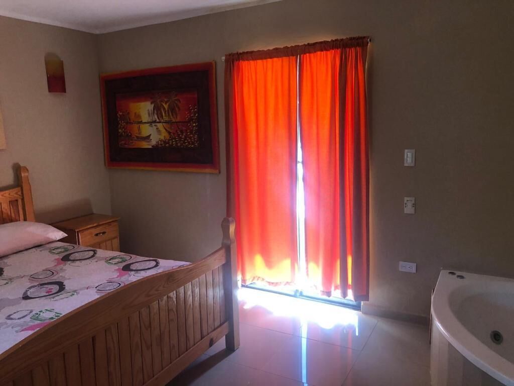 Quarto com cama, mesa de cabeceira, quadro, janela com cortinas laranja e banheira.