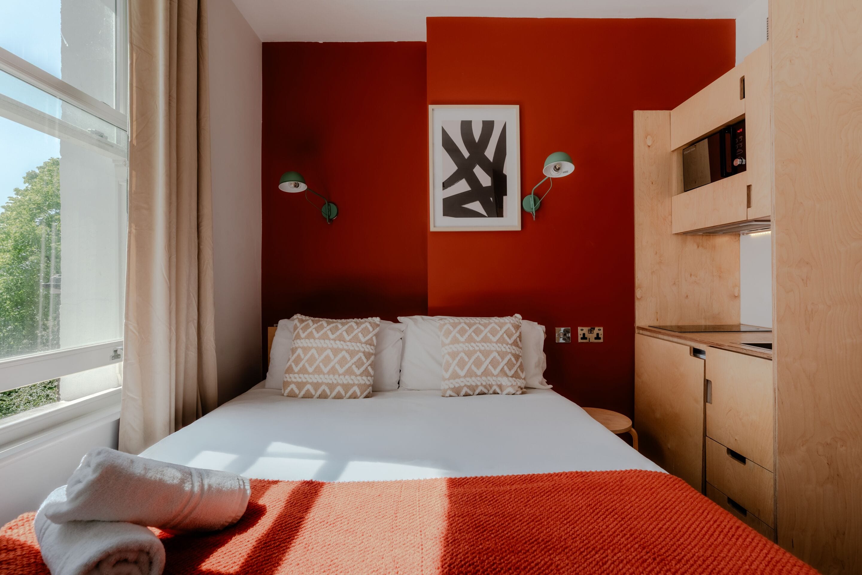 Quarto de hotel com cama, parede de destaque vermelha, obra de arte abstrata emoldurada e cozinha compacta.
