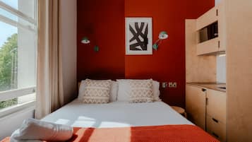 Quarto de hotel com cama, parede de destaque vermelha, obra de arte abstrata emoldurada e cozinha compacta.