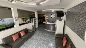 Um saguão de hotel com uma mesa de recepção, dois sofás com almofadas, uma televisão na parede e um papel de parede estampado.