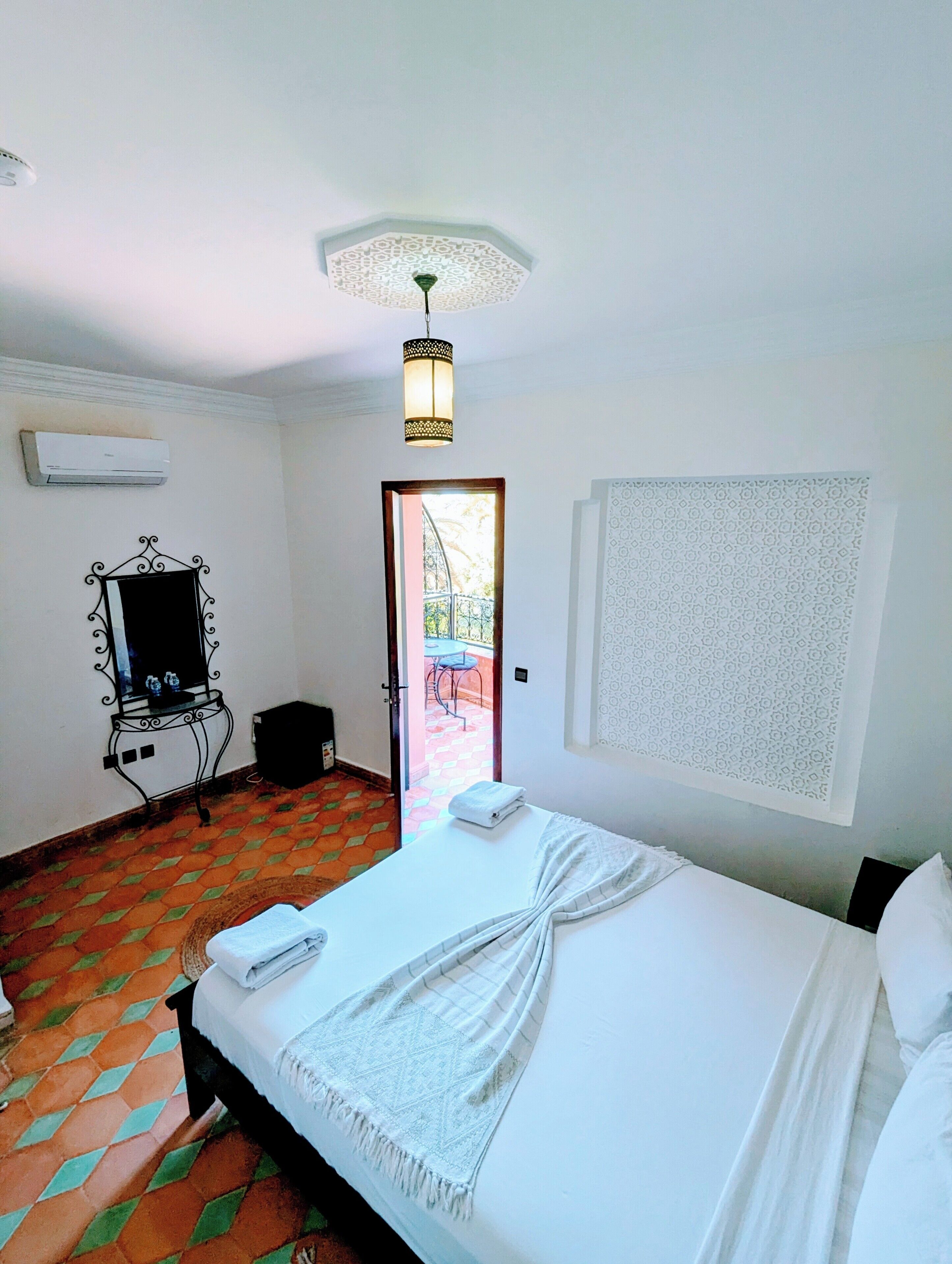 Quarto de hotel com cama, luminária no teto, espelho e porta para área externa.