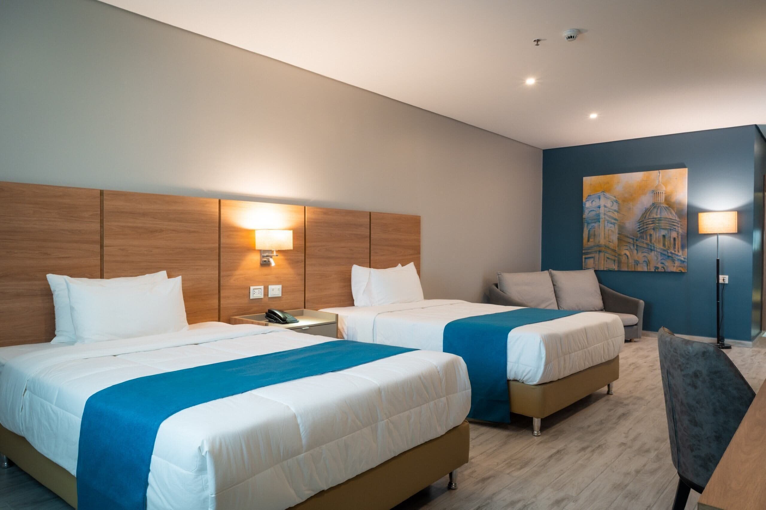 Quarto de hotel com duas camas, uma escrivaninha, uma cadeira e um quadro na parede.