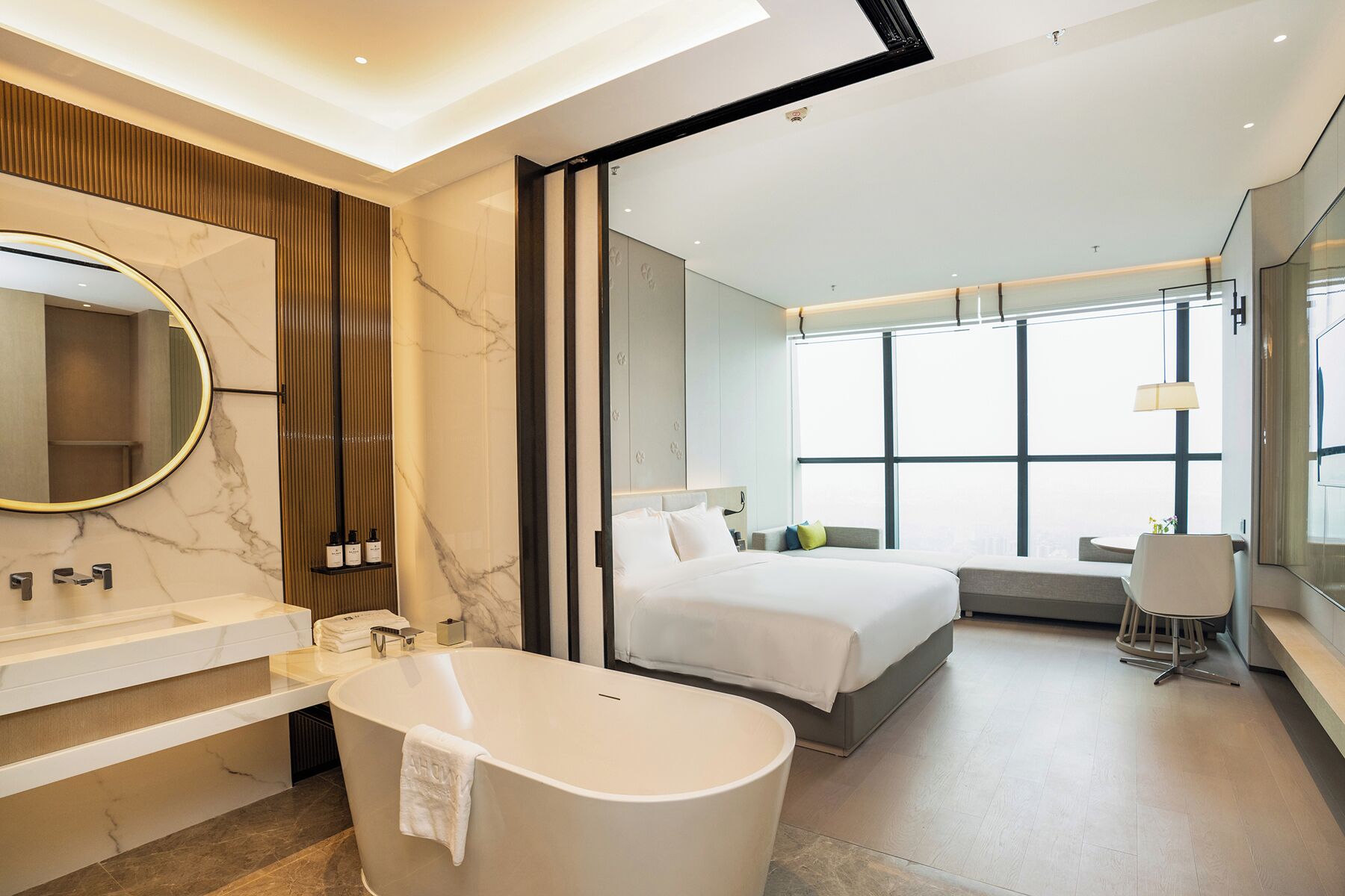Quarto de hotel moderno com uma cama grande, área de estar separada, banheira e uma janela ampla com vista para a cidade.