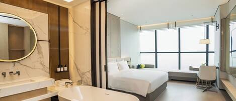Quarto de hotel moderno com uma cama grande, área de estar separada, banheira e uma janela ampla com vista para a cidade.