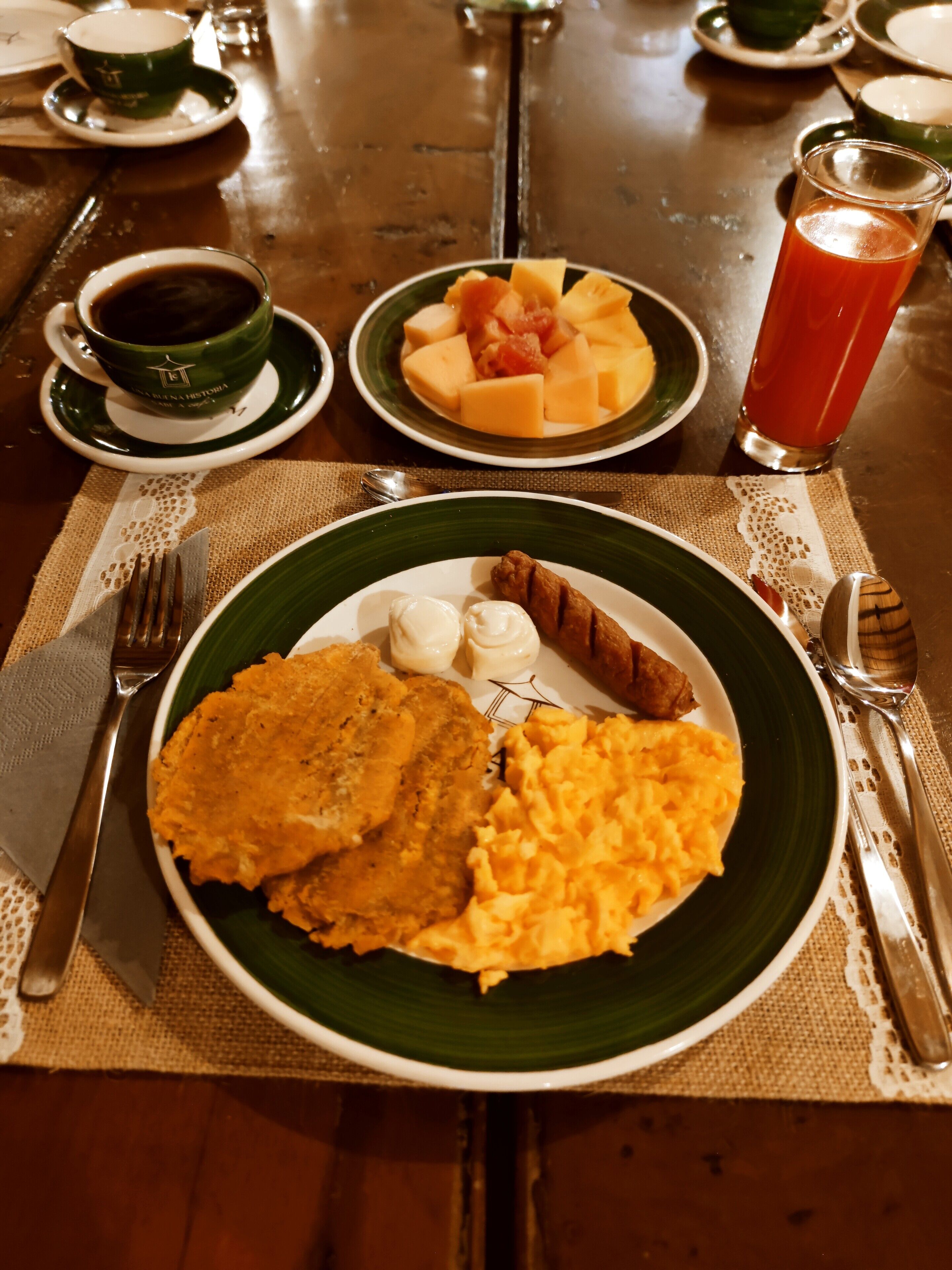 Prato de café da manhã com escalopado, ovos mexidos e salsicha, acompanhado de uma xícara de café verde e uma tábua de frutas.