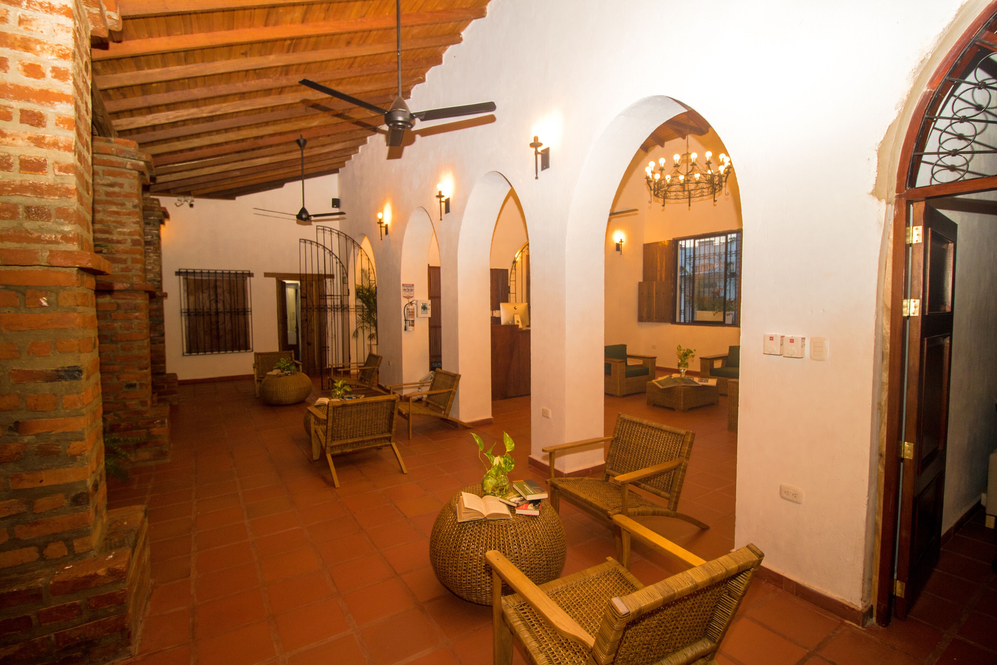 Um hall espaçoso com pé direito alto, vigas de madeira e um ventilador de teto. Há várias cadeiras de vime e um banquinho redondo com uma planta e livros. As paredes possuem portas em arco e uma grande janela com vista.