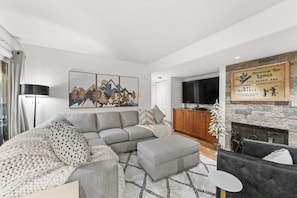 Uma sala de estar moderna com um sofá seccional cinza, um banquinho cinza e uma poltrona preta. Há uma TV de tela plana na parede, uma lareira de pedra e uma placa emoldurada sobre a lareira.