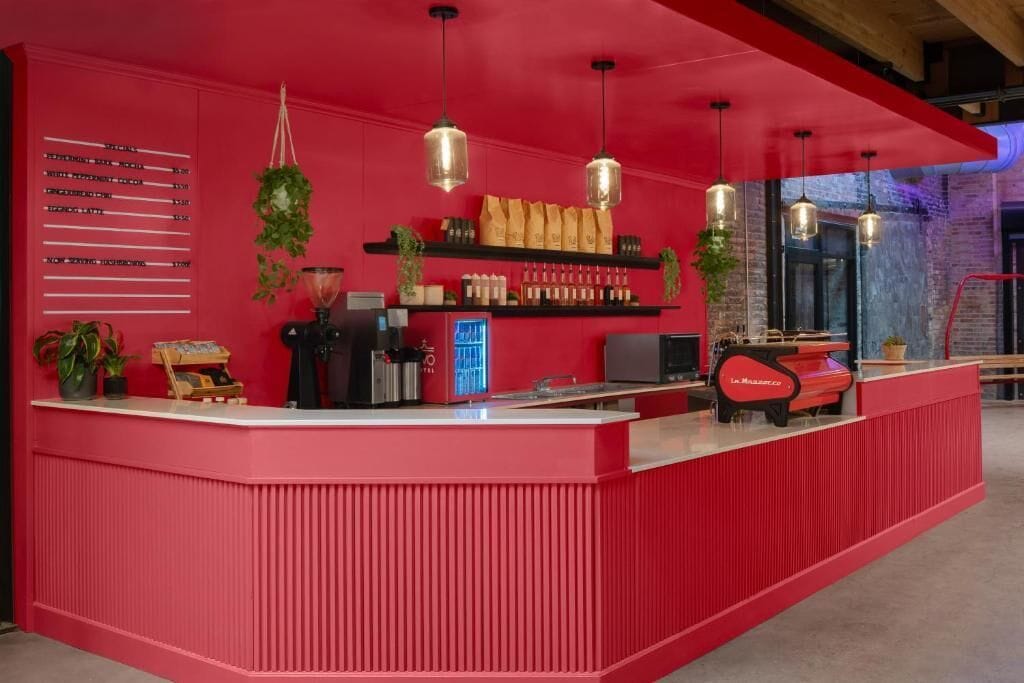 Um balcão de recepção vermelho com plantas pendentes, uma máquina de café e uma prateleira com sacolas e garrafas.