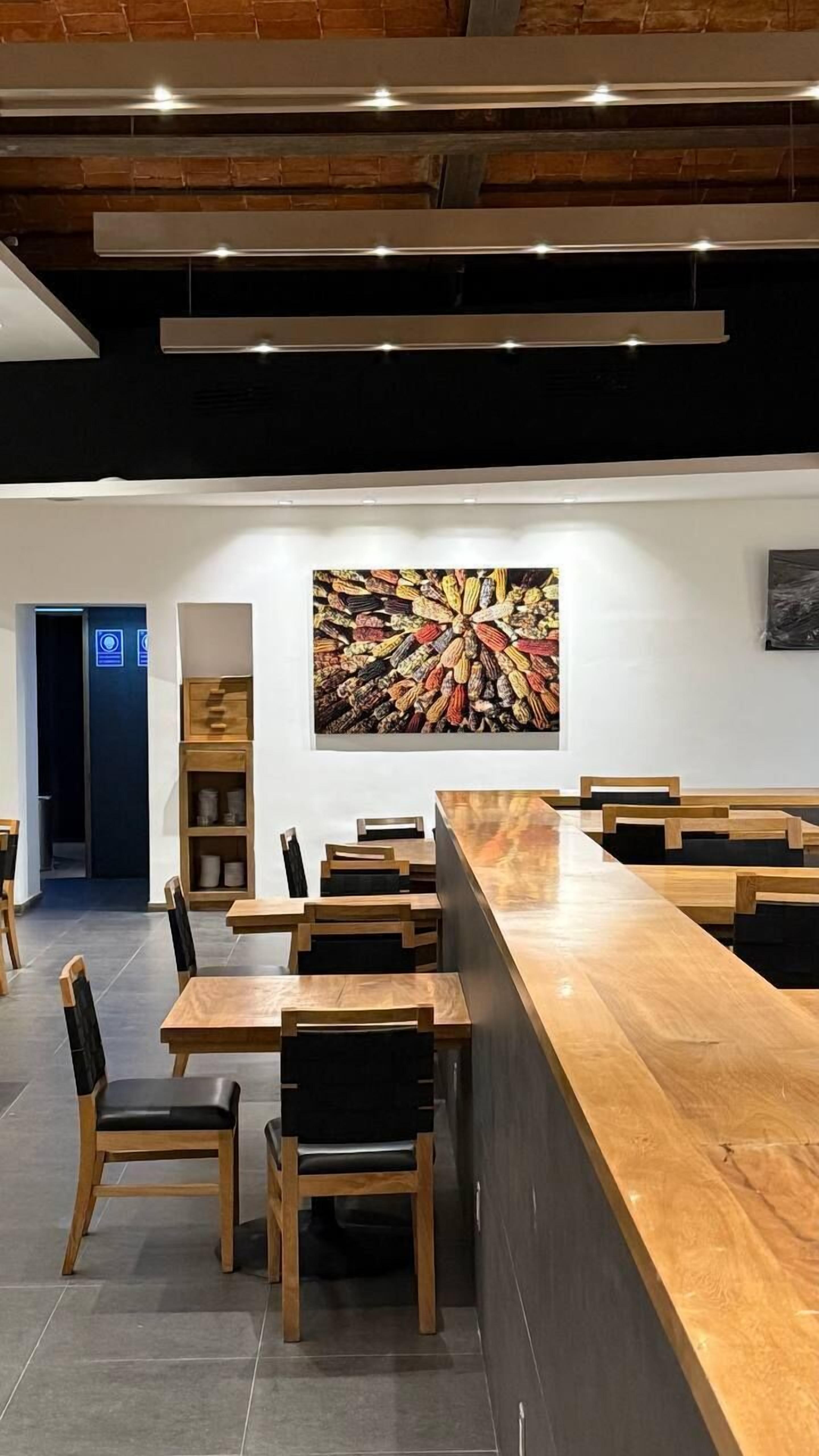 Interior moderno de restaurante com mesas e cadeiras de madeira, uma obra de arte central e teto preto.