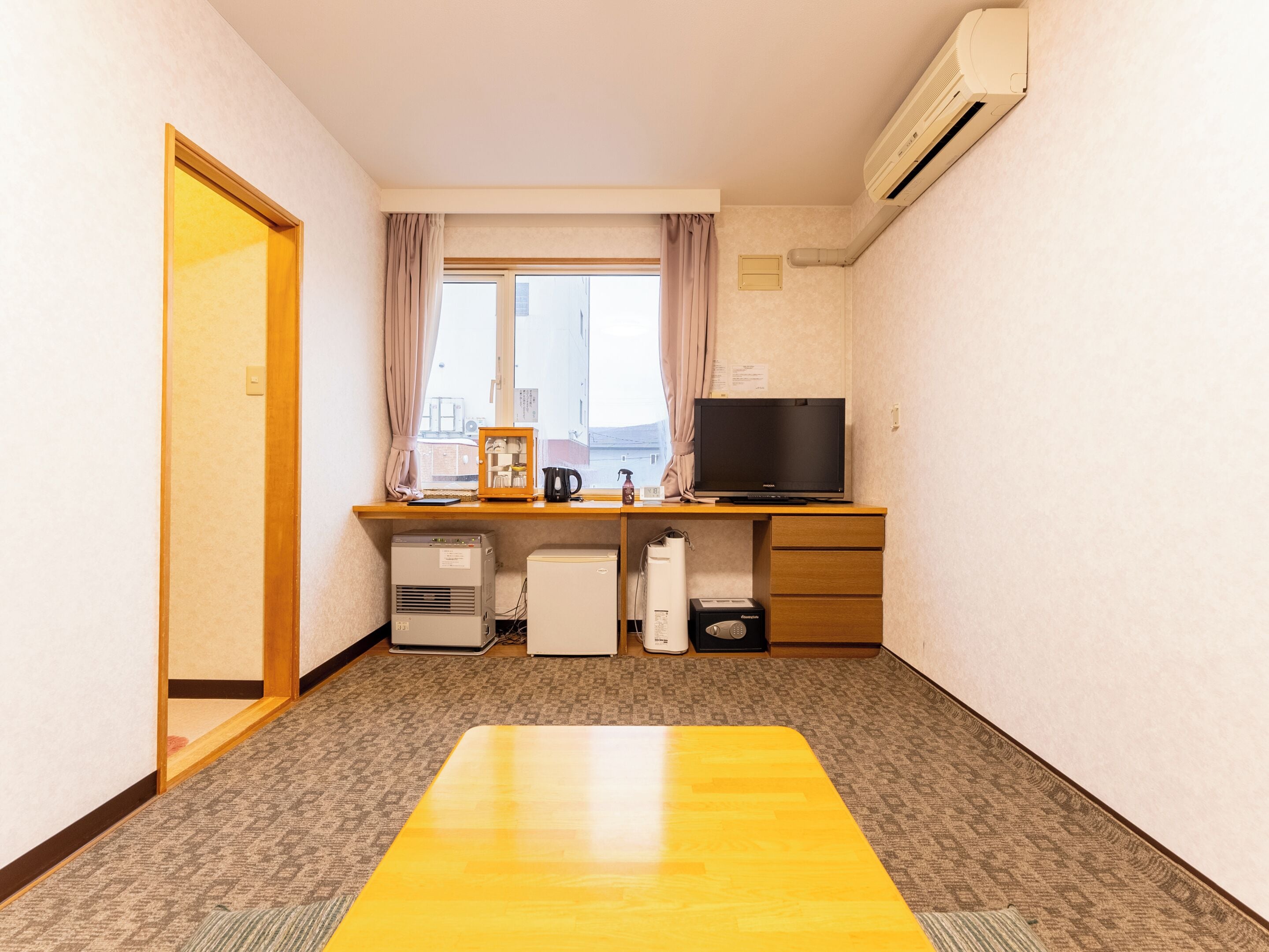 Quarto de hotel pequeno com escrivaninha de madeira, televisão, mini-frigorífico, cafeteira e janela com cortinas.