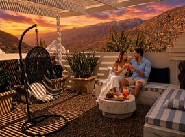 Um casal sentado numa varanda com vista para montanhas, uma cadeira suspensa e uma mesa com petiscos.