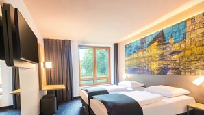Quarto de hotel com duas camas, uma TV de tela plana na parede, uma janela com vista para áreas verdes e uma pintura vibrante de paisagem urbana na parede.