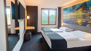 Quarto de hotel com cama, escrivaninha, cadeira, lixeira, janela e um quadro na parede.