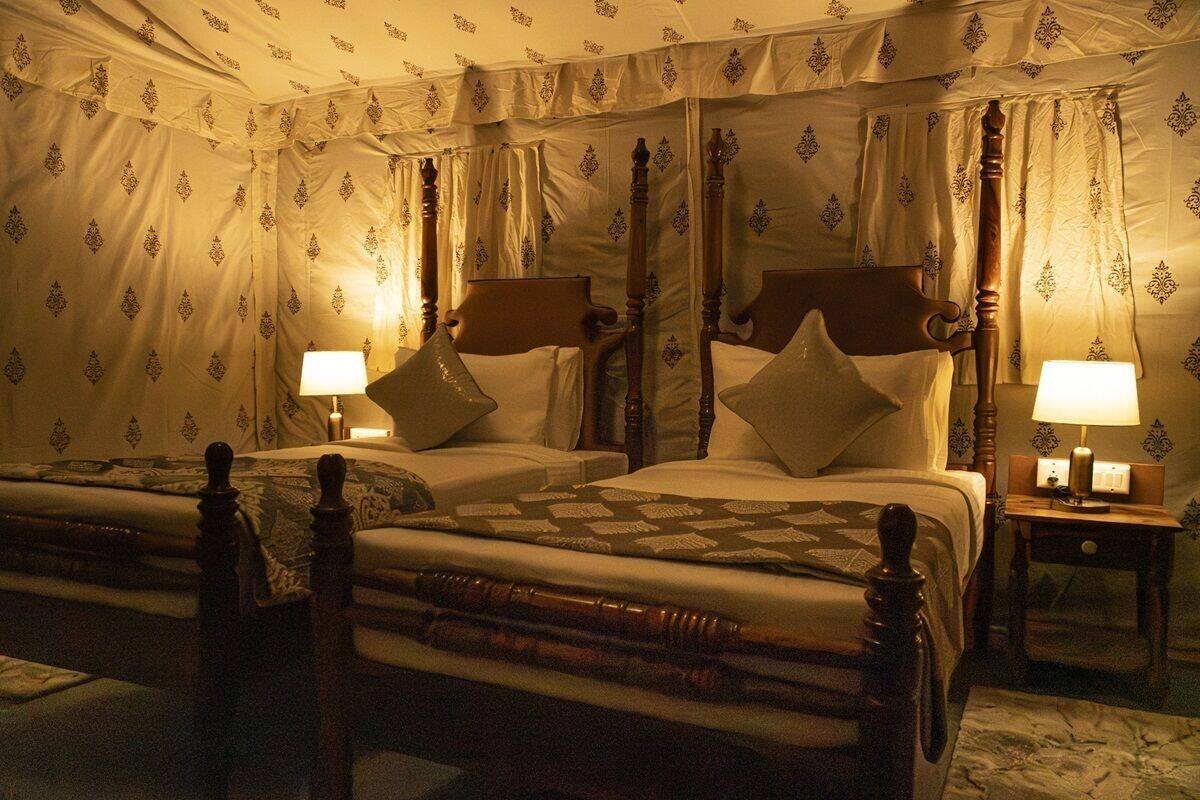 Um quarto tradicional com duas camas de madeira, cada uma com encosto, e uma mesinha de cabeceira com abajur. O quarto possui papel de parede estampado e cortinas.