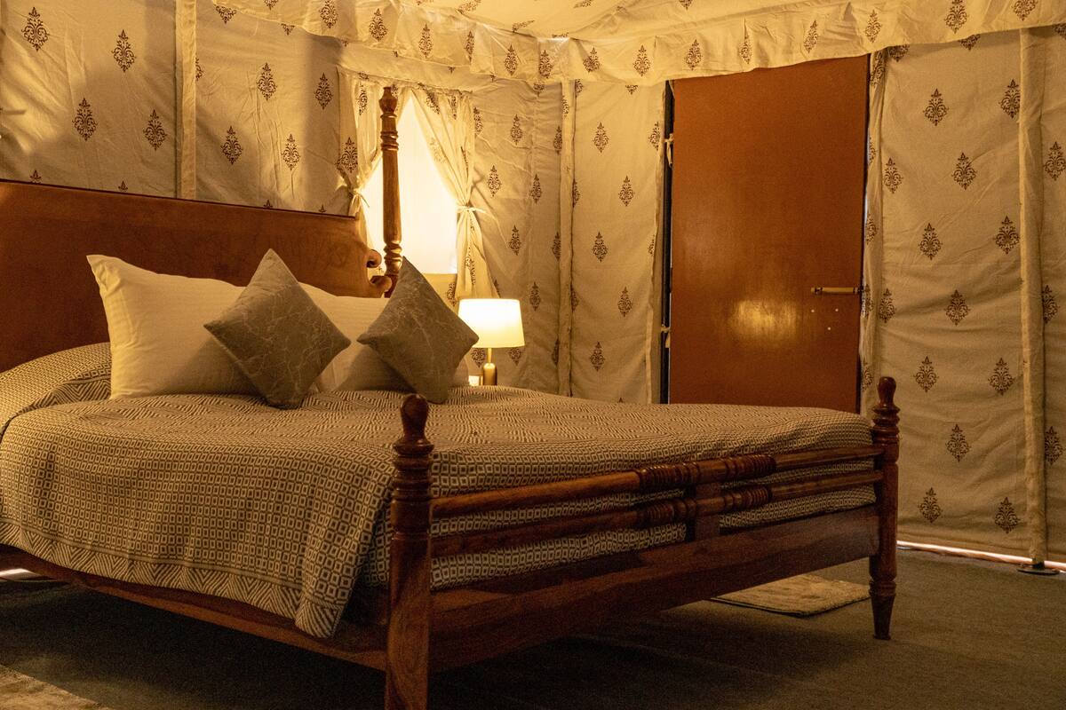 Uma cama de madeira com dossel, com edredão e almofadas estampadas, uma luminária de cabeceira e uma porta com cortinas padronadas.