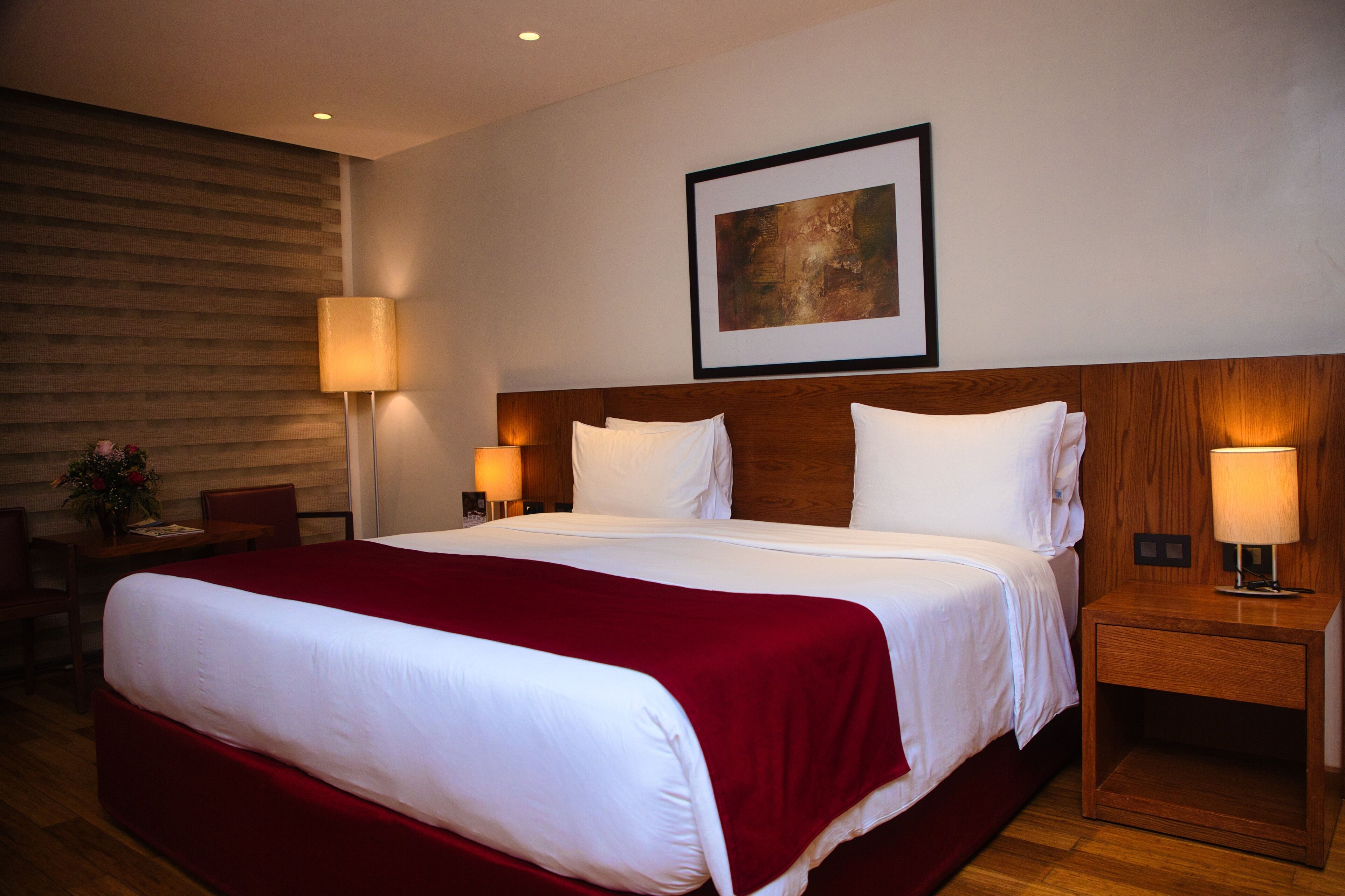 Quarto de hotel com uma cama grande, duas mesas de cabeceira com luminárias, cabeceira de madeira, um quadro na parede e piso de madeira.