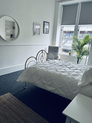 Quarto de hotel bem arrumado, com cama, uma mesinha, uma cadeira, um espelho e uma janela com vista para edifícios.