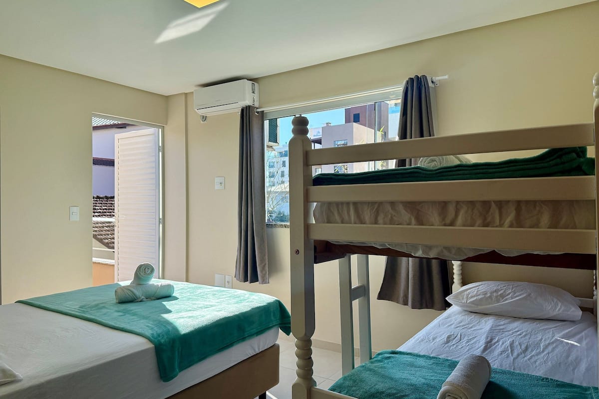 Quarto com duas camas, uma com cobertor verde e outra com roupa de cama branca. Há uma janela com cortinas, um ar condicionado na parede e uma porta que dá para o exterior.